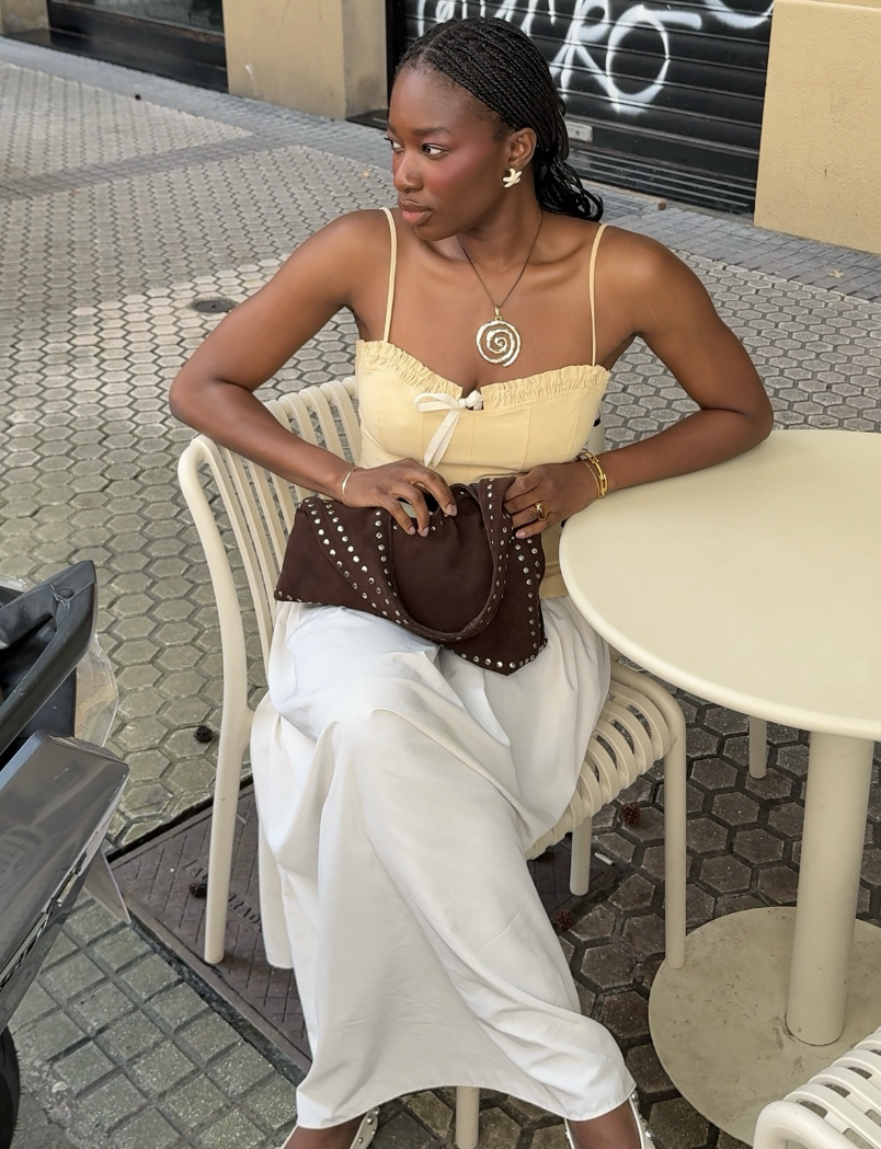 Woman wearing a yellow spaghetti strap top with ruffle detail%2C white flowy skirt%2C white studded flats%2C and holding a brown studded handbag while sitting at an outdoor cafe table